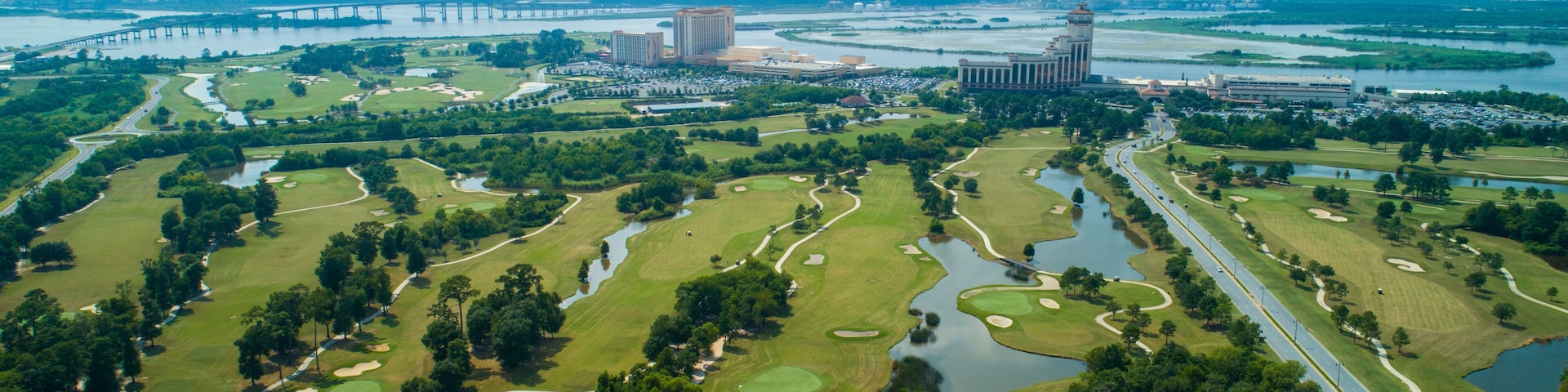 Um campo de golfe com obstáculos de água, uma ponte e um horizonte urbano ao fundo.