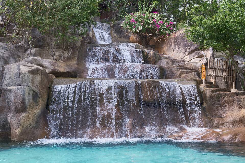 Uma cascata em vários níveis deságua em uma piscina azul-turquesa, cercada por grandes rochas e vegetação exuberante.