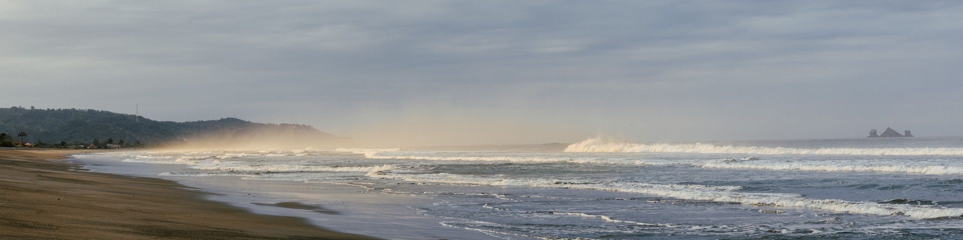 Uma praia com ondas quebrando na costa, uma cadeia montanhosa ao longe e um céu nublado.