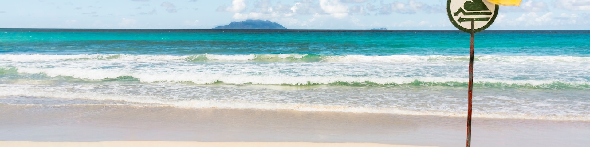 Uma praia com uma bandeira em um mastro, oceano turquesa e uma ilha ao longe.
