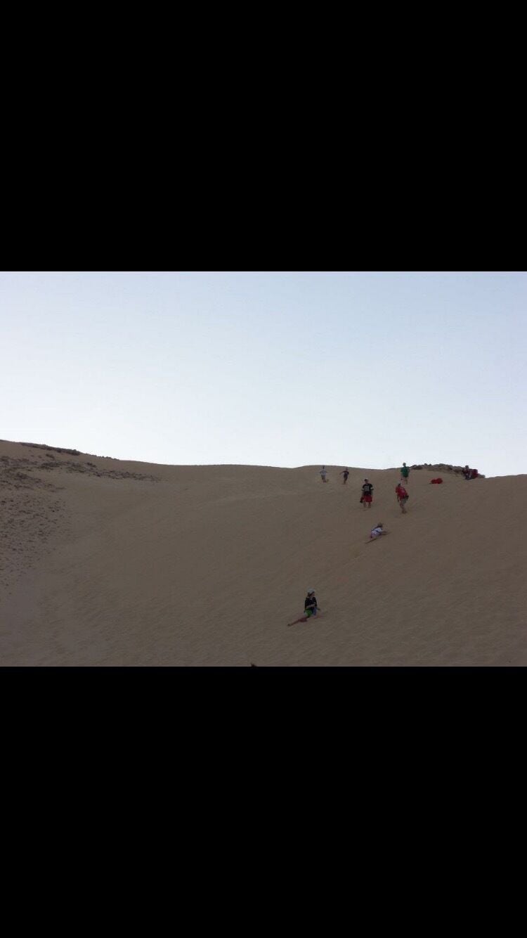 Paisagem desértica arenosa com um grupo de pessoas escalando uma duna de areia.