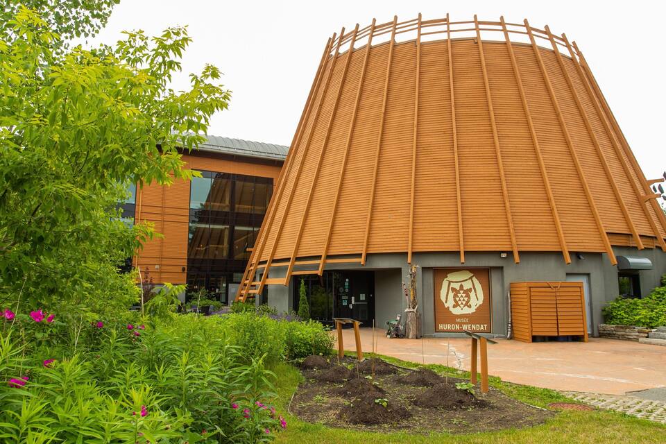 Um museu com um telhado de madeira cónico e uma placa com a inscrição 'MUSEE HURON-WENDAT'.