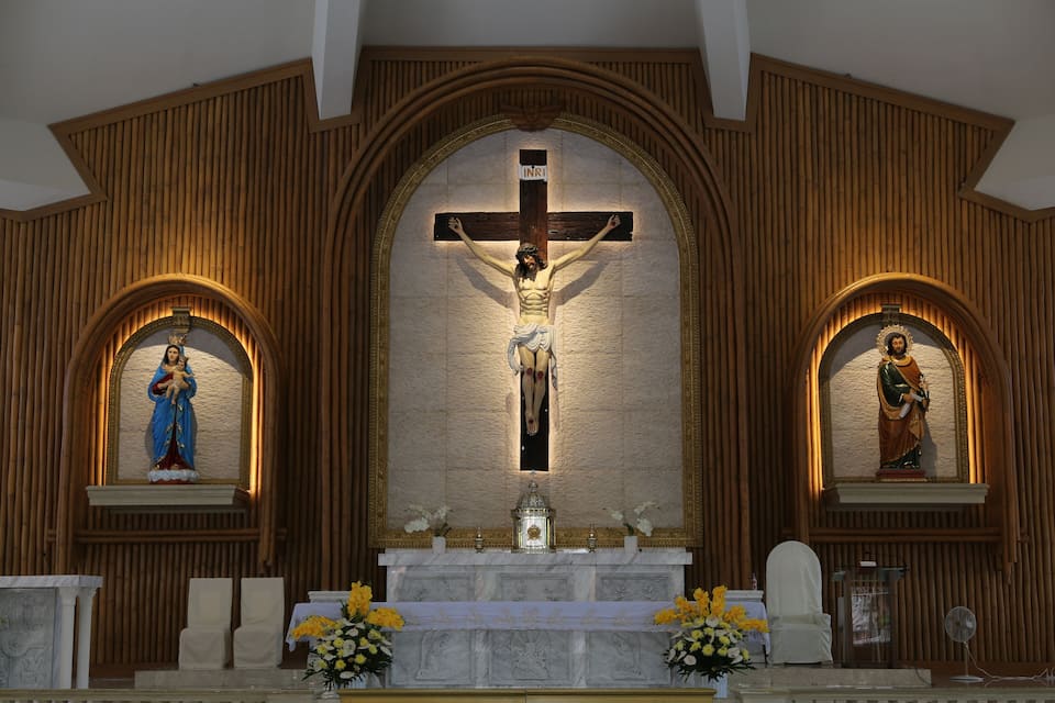 Interior de igreja com crucifixos, duas estátuas, altar e arranjos florais.