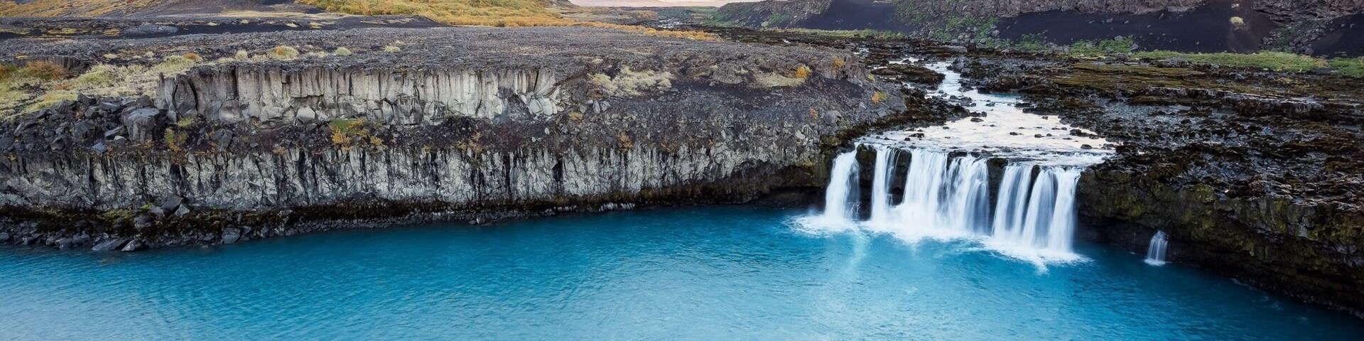 Uma cascata deságua em um lago azul, cercado por penhascos rochosos e uma montanha ao fundo.
