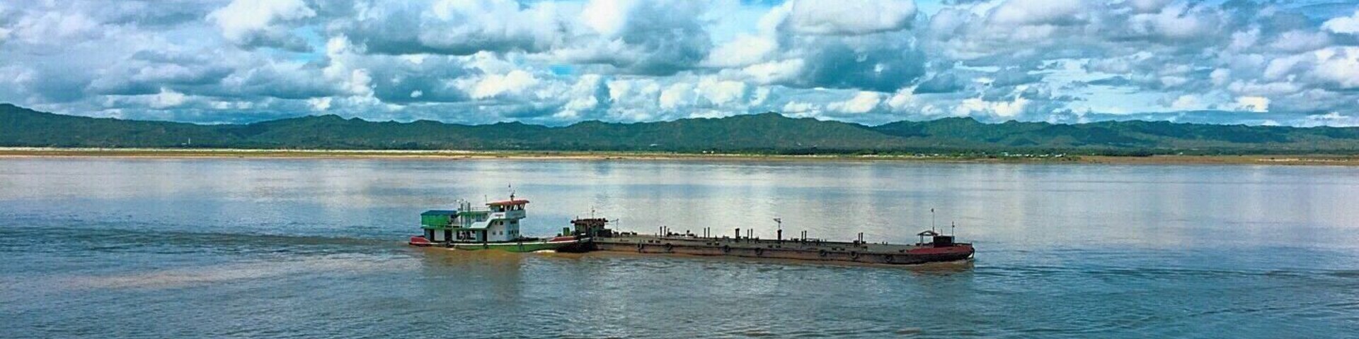 Um grande navio de carga em águas calmas, com um pequeno rebocador nas proximidades. Ao fundo, montanhas e um céu parcialmente nublado.