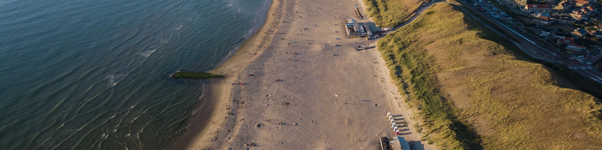 Vista aérea de uma área costeira com uma praia arenosa, uma estrada paralela à costa e um conjunto de edifícios à direita.