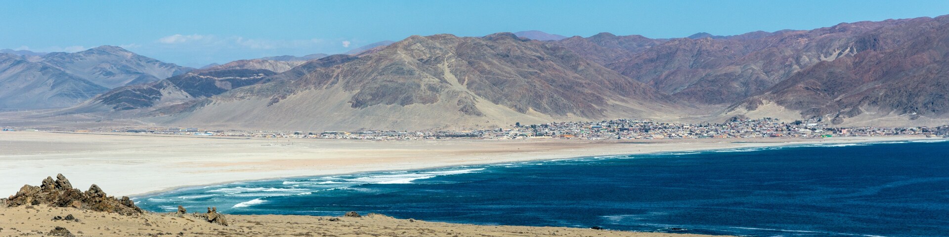 Paisagem costeira com praia arenosa, afloramento rochoso e uma cidade aninhada entre montanhas.