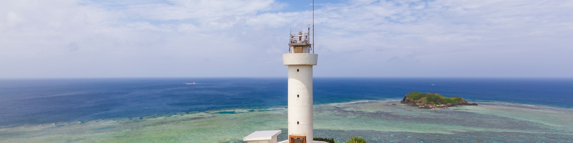 Um farol numa colina gramada com vista para um mar azul e límpido, com uma pequena ilha ao longe.