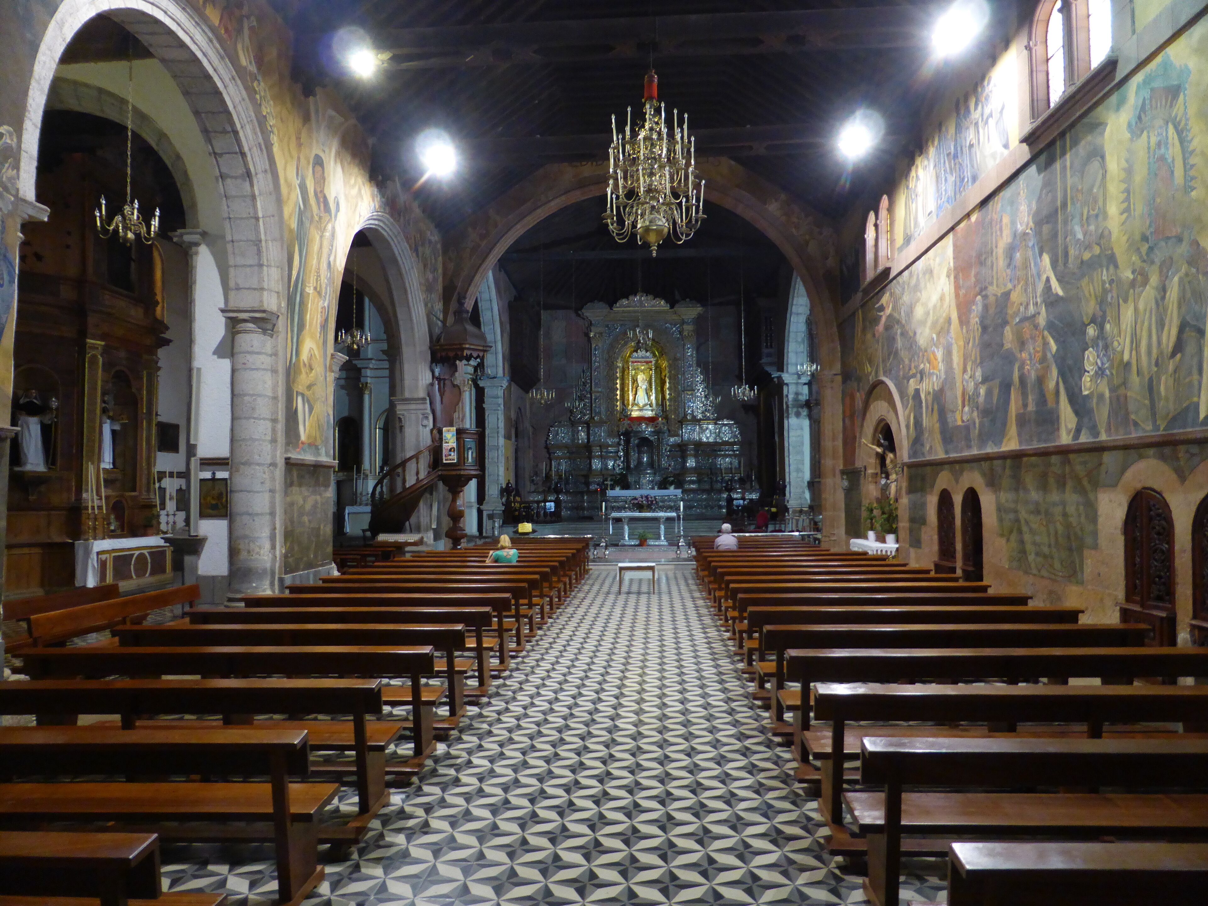 Interior de uma igreja com bancos de madeira, piso estampado e um grande altar.