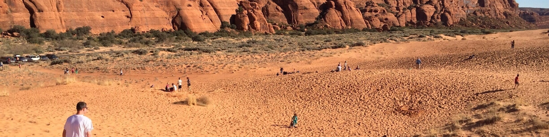 Paisagem desértica com formações rochosas avermelhadas e pessoas caminhando e sentadas na areia.
