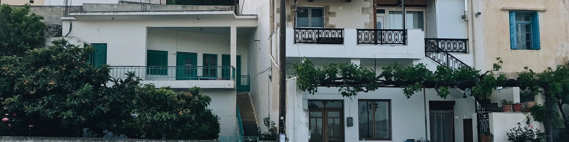 Fileira de edifícios brancos com varandas e persianas verdes, rodeados por árvores e plantas em vasos.