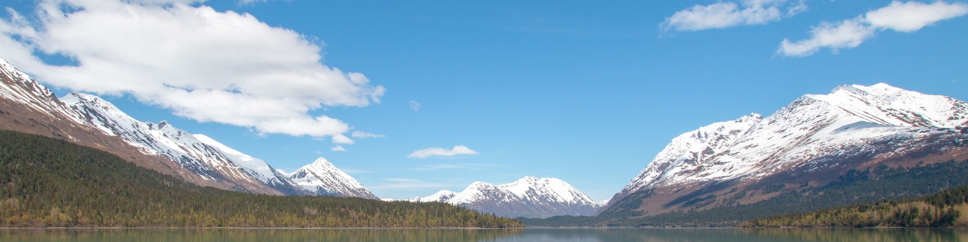 Um lago calmo refletindo montanhas nevadas, céu azul límpido e um vale florestado.