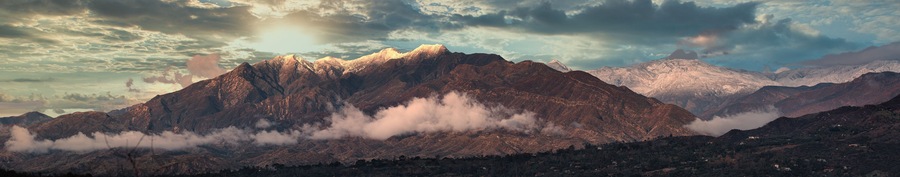Uma cordilheira com picos nevados, nuvens e um vale abaixo.