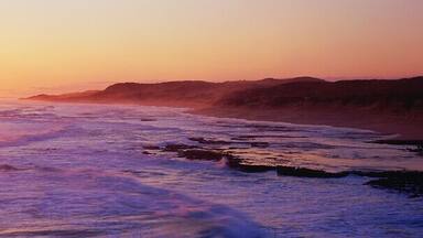 Um pôr do sol sobre uma praia com ondas e uma linha costeira distante.