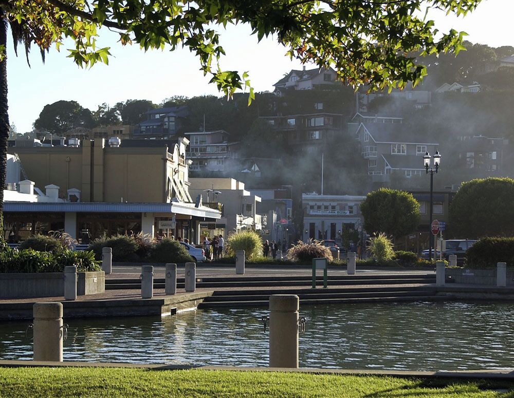 Um parque com uma fonte, um edifício com um letreiro e uma colina com casas ao fundo.