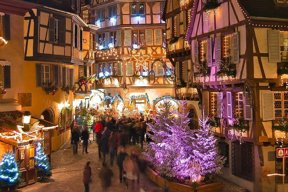 Ruada de paralelepípedos estreita, ladeada por edifícios tradicionais em estilo de enxaimel, decorados com luzes festivas e enfeites de Natal.