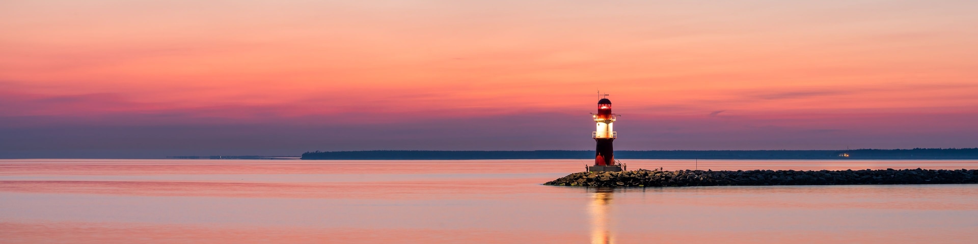 Um farol sobre um afloramento rochoso ao pôr do sol, com águas calmas refletindo o céu.