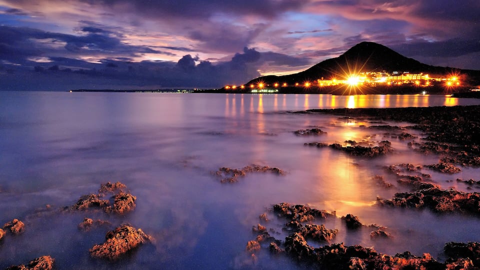 Paisagem costeira ao crepúsculo, com montanha, águas calmas e edifícios iluminados refletindo na água.