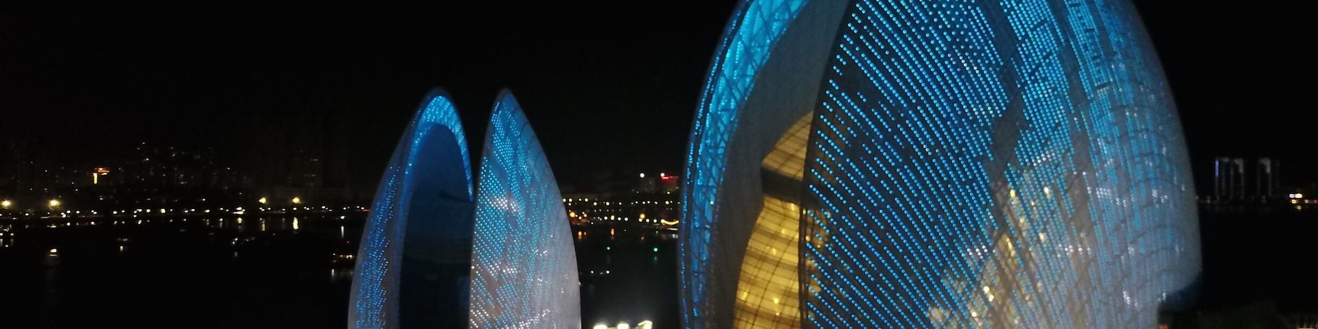 Vista noturna de uma estrutura arquitetônica moderna com padrões azuis iluminados, uma ponte e um horizonte urbano ao fundo.