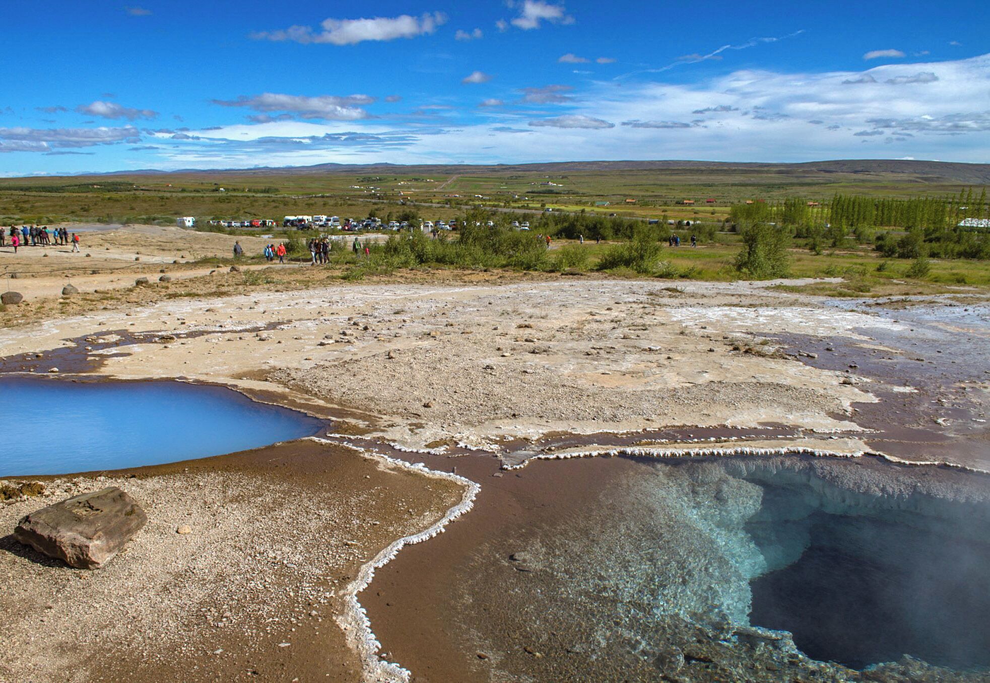 Uma área geotérmica com nascentes termais, um pequeno lago e um povoado distante.