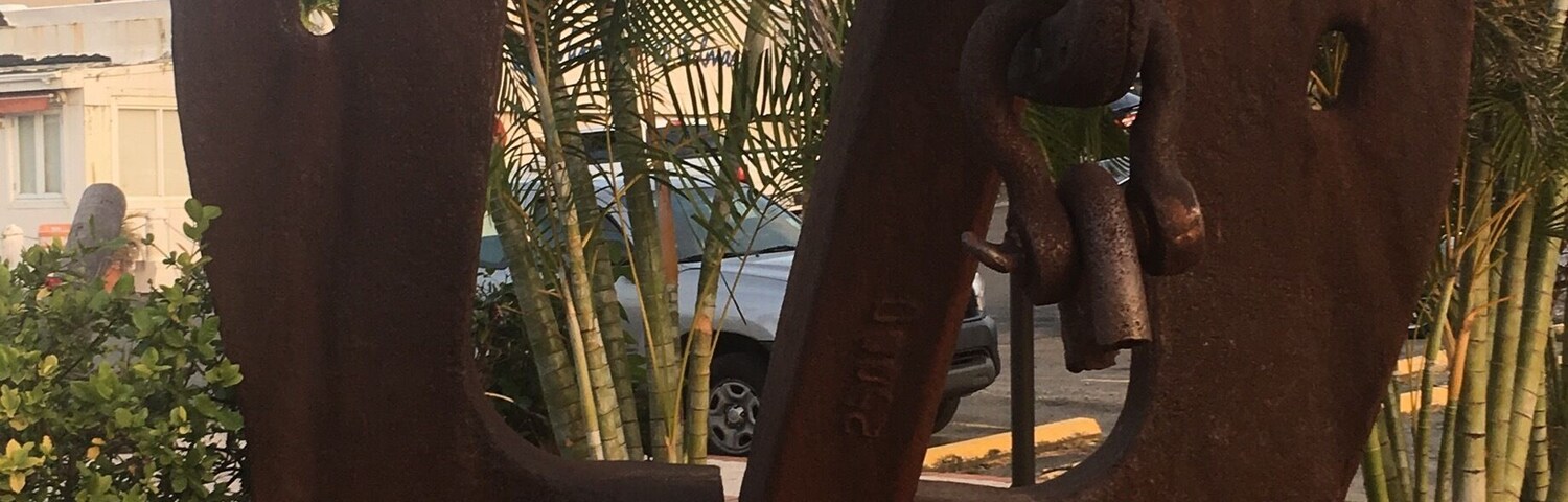 Uma escultura de metal enferrujado com um buraco no centro, rodeada por palmeiras e um carro ao fundo.