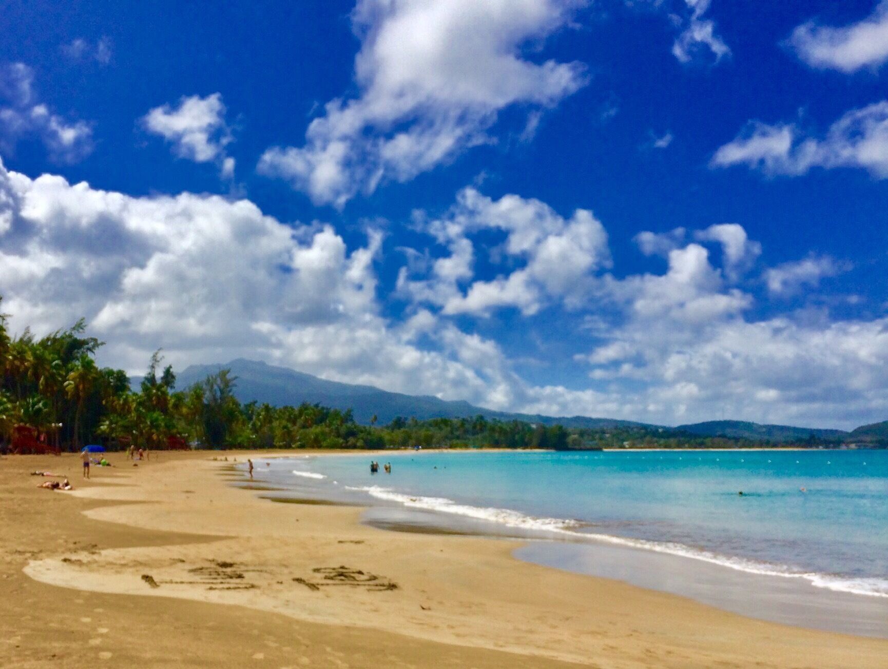 Uma praia com águas turquesa cristalinas, palmeiras e montanhas ao fundo.