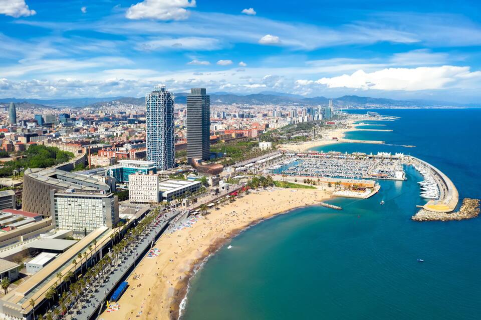 Paisagem costeira com uma praia extensa, um porto de iates e edifícios altos.
