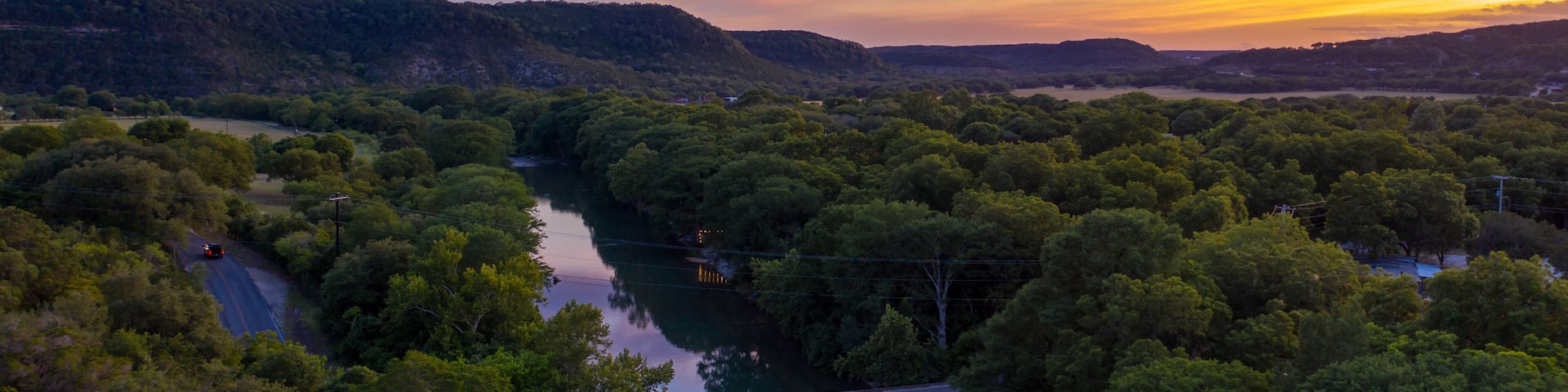 Um rio serpenteia por um vale florestado, com colinas ao fundo e uma ponte cruzando o rio.
