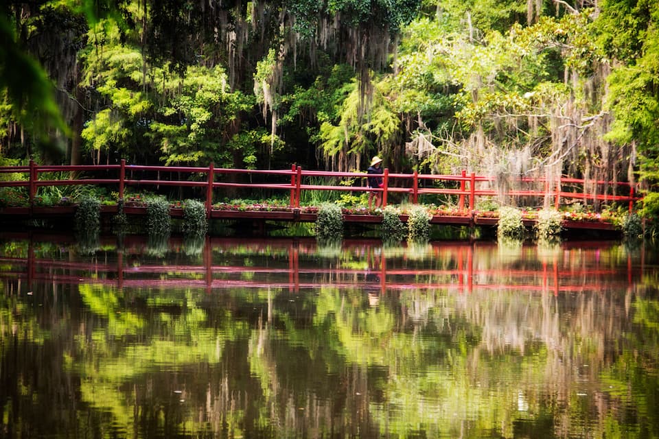 Uma ponte vermelha sobre um lago tranquilo, rodeada por vegetação exuberante e árvores.