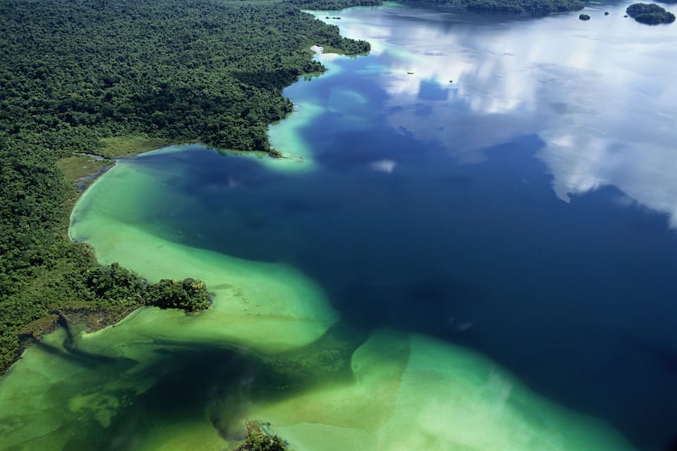 Vista aérea de uma área costeira com águas azul-claras, bancos de areia esverdeados e terreno florestado denso.