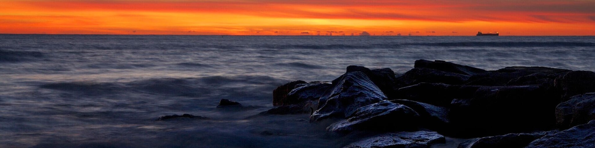 Um pôr do sol sobre o oceano, com rochas em primeiro plano.
