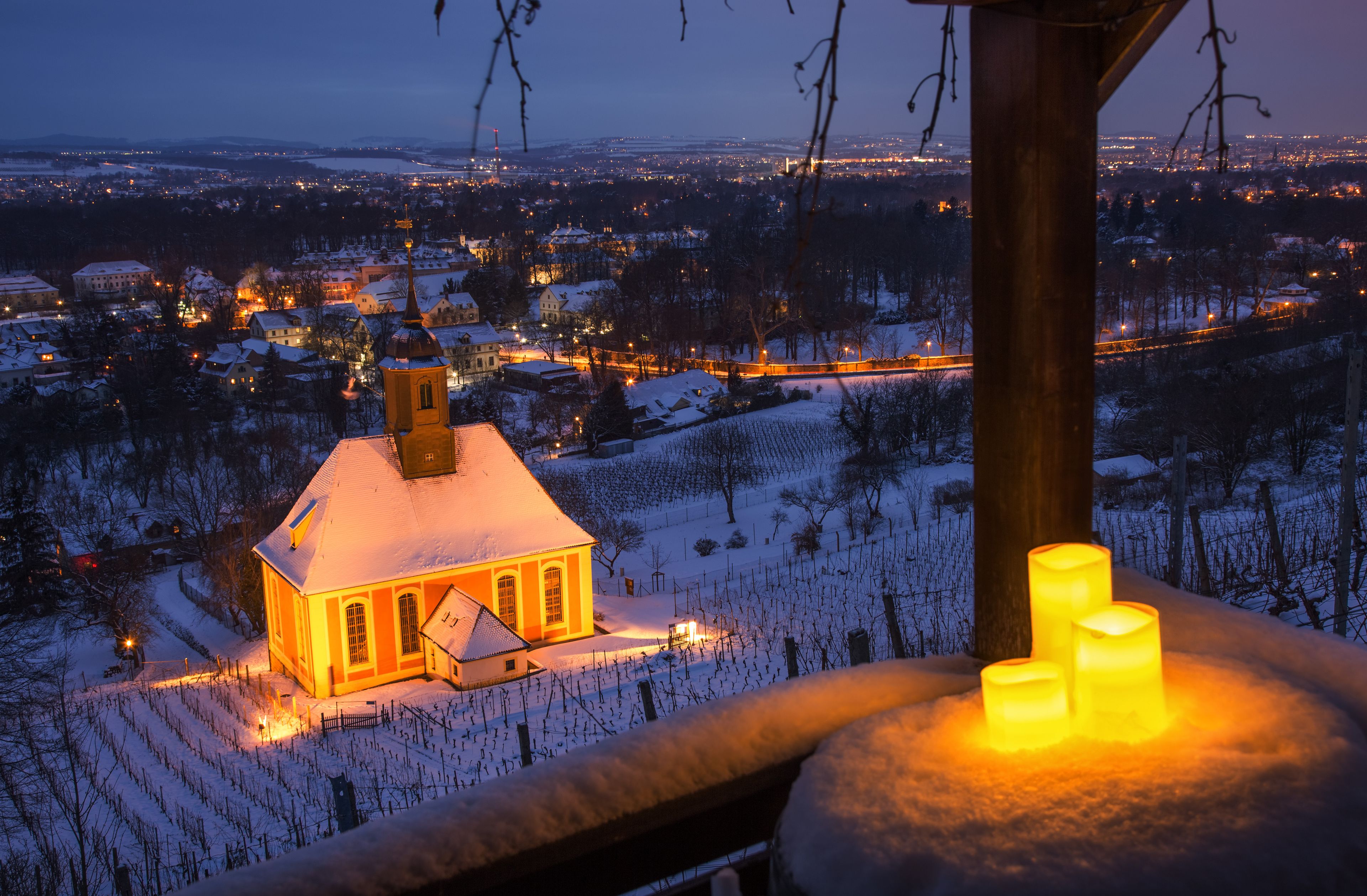 Paisagem coberta de neve com uma igreja iluminada e uma cidade distante ao crepúsculo.