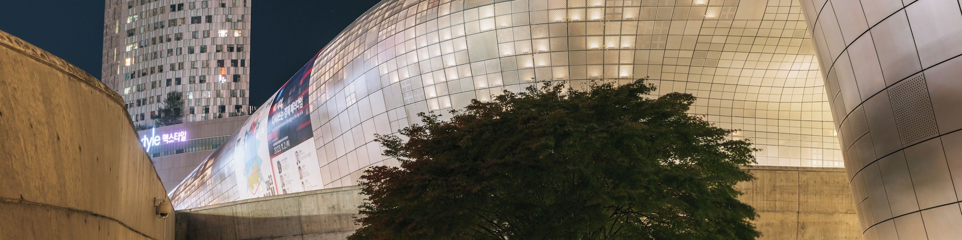 Edifício moderno com fachada de vidro curva e uma árvore em primeiro plano.