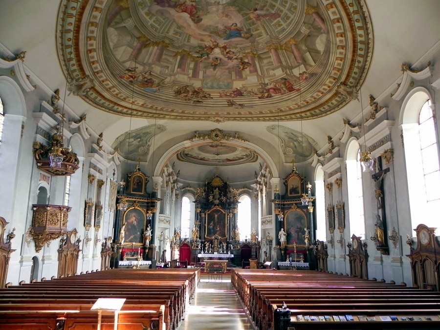 Interior de igreja ornamentado, com altares, tetos imponentes, afrescos detalhados e fileiras de bancos de madeira.