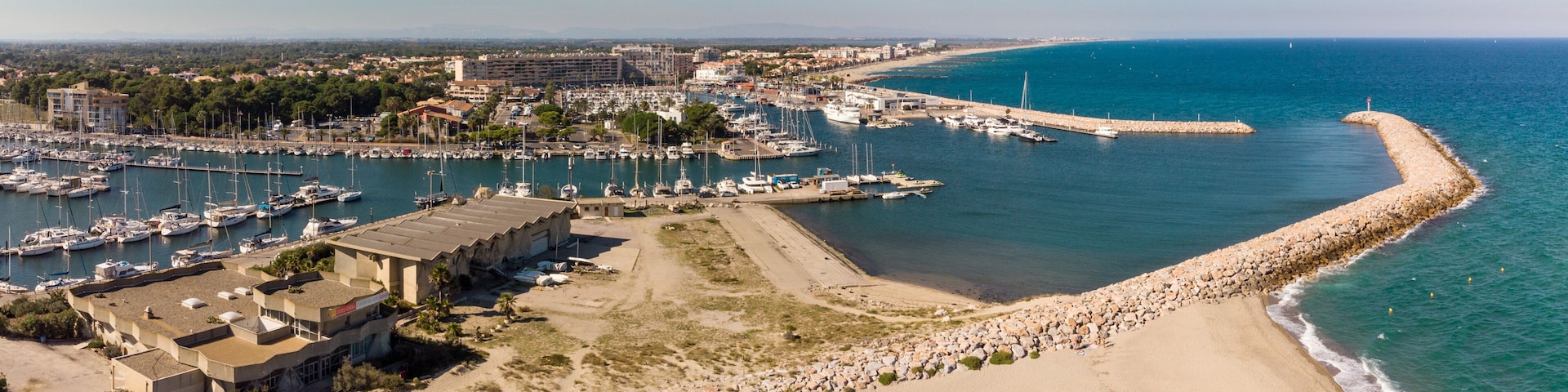 Uma marina costeira com barcos, uma praia de areia e um molhe rochoso.