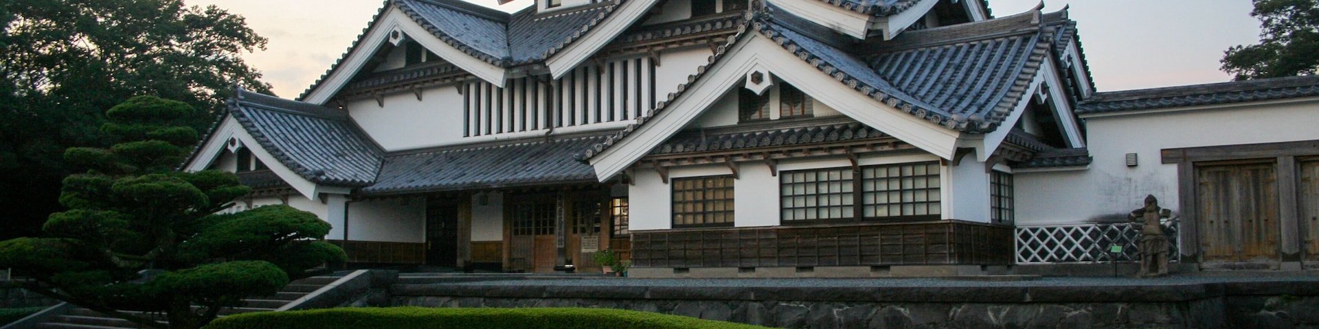 Um castelo japonês tradicional com vários níveis, varandas de madeira e um jardim bem cuidado, com arbustos aparados e um lago.