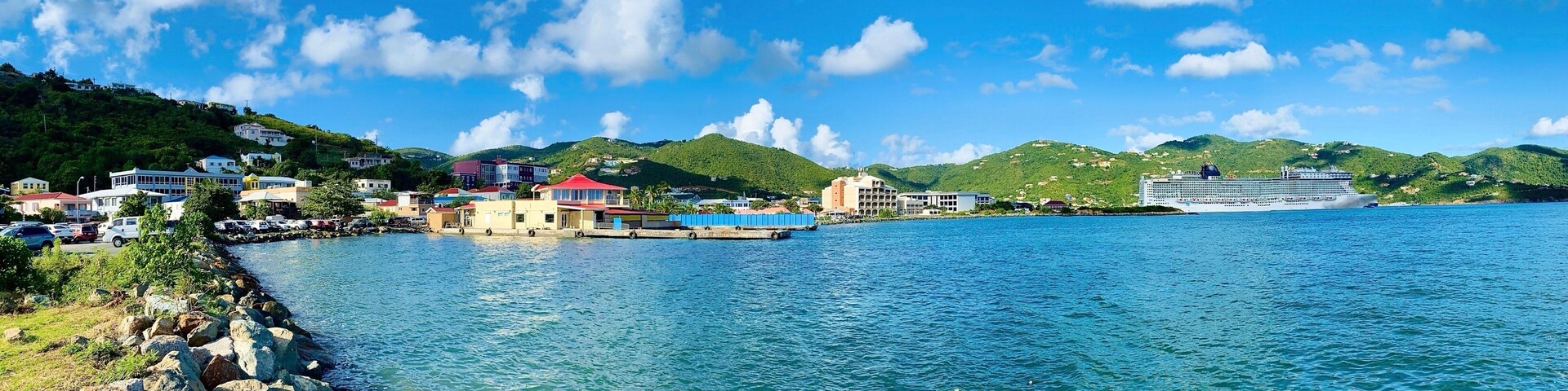 Uma cidade costeira com um cais, um navio de cruzeiro e colinas verdes ao fundo.