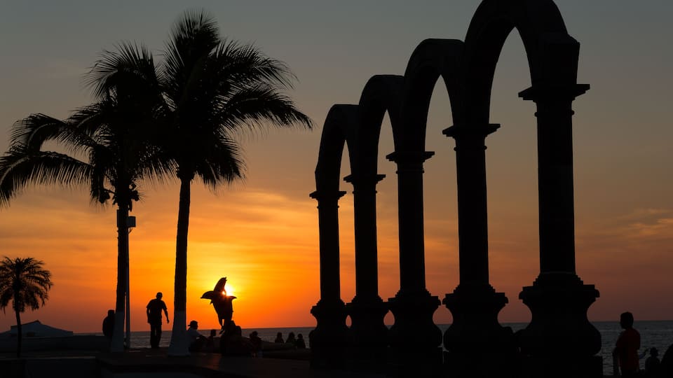 Pôr do sol com silhuetas de palmeiras e colunas clássicas.