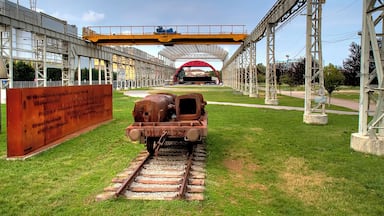 Trilhos de ferro com uma locomotiva de trem antiga, uma grande estrutura metálica acima e uma placa com texto.