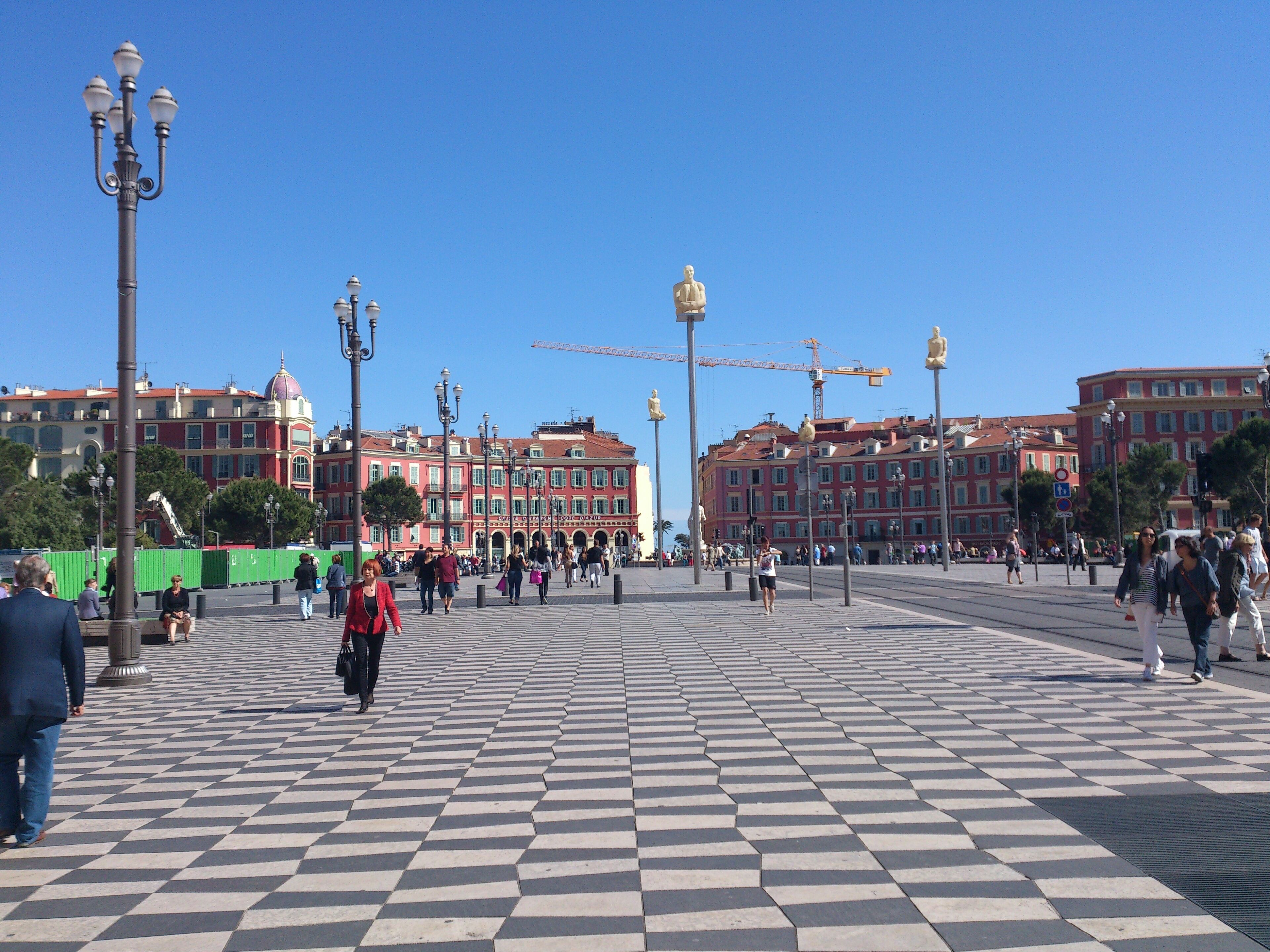 Uma praça com piso xadrez, pessoas caminhando e edifícios históricos ao fundo.