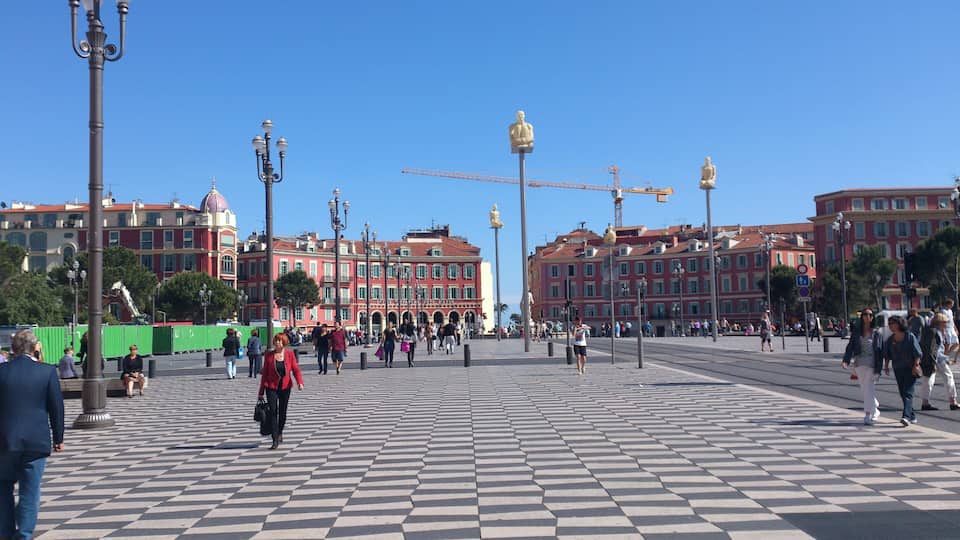 Uma praça com piso xadrez, pessoas caminhando e edifícios históricos ao fundo.