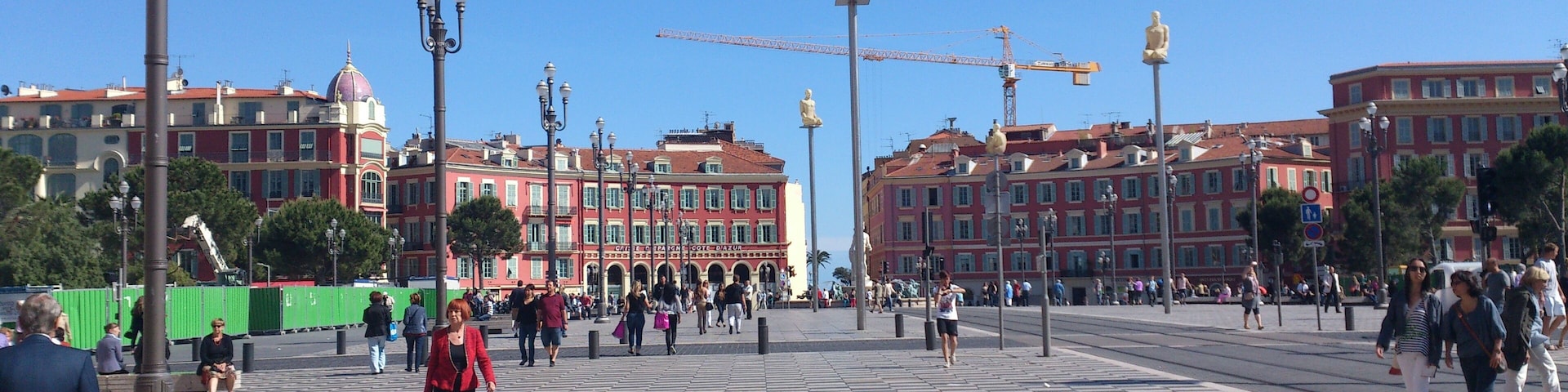 Uma praça com piso xadrez, pessoas caminhando e edifícios históricos ao fundo.