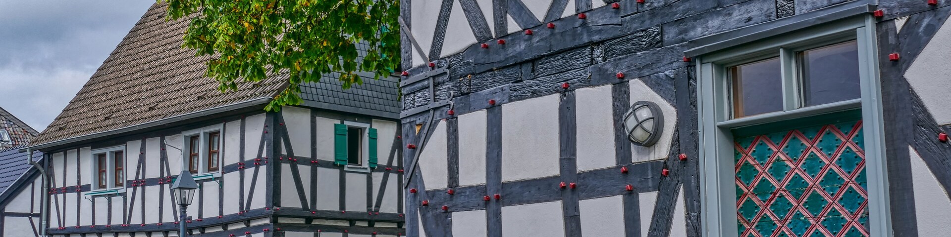 Uma casa tradicional em estilo de enxaimel com porta e venezianas verdes, rodeada por ruas de calçada e uma árvore.