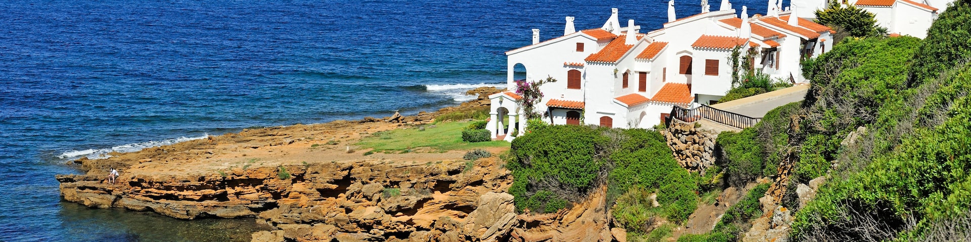 Uma vila costeira com edifícios brancos e telhados vermelhos, falésias rochosas e o mar.