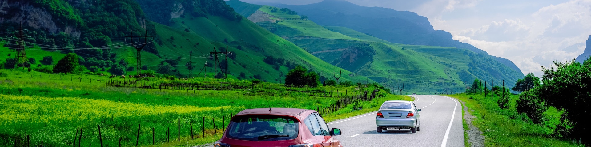 Um carro vermelho dirigindo em uma estrada, com campos verdes e montanhas ao fundo.