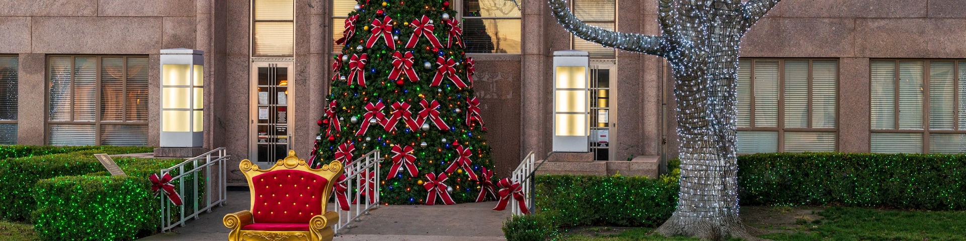 Um edifício com uma grande árvore de Natal decorada com fitas e luzes vermelhas e brancas, uma cadeira dourada e duas guirlandas nas portas.