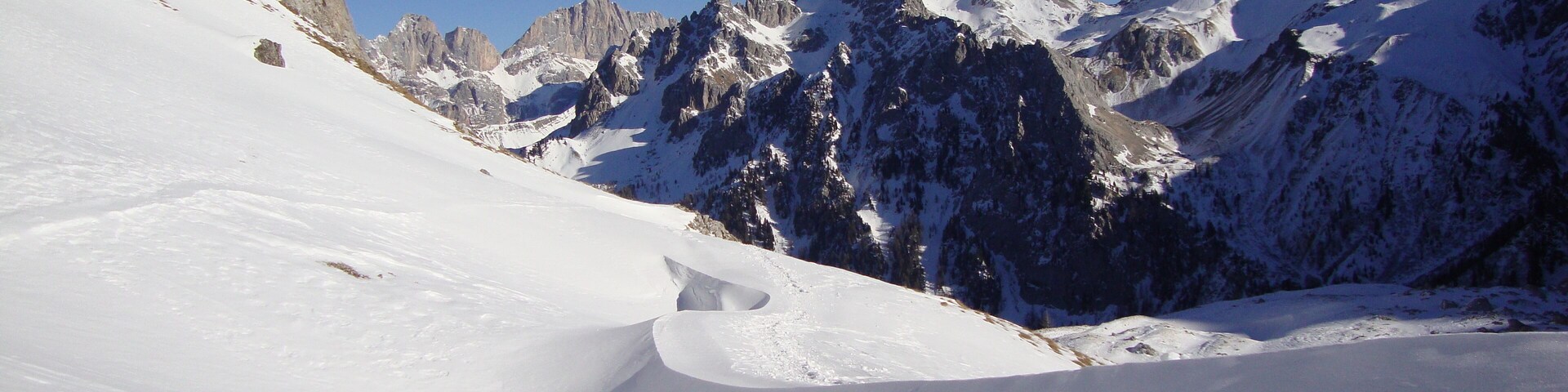 Encosta de montanha coberta de neve, com afloramentos rochosos e um céu azul límpido.
