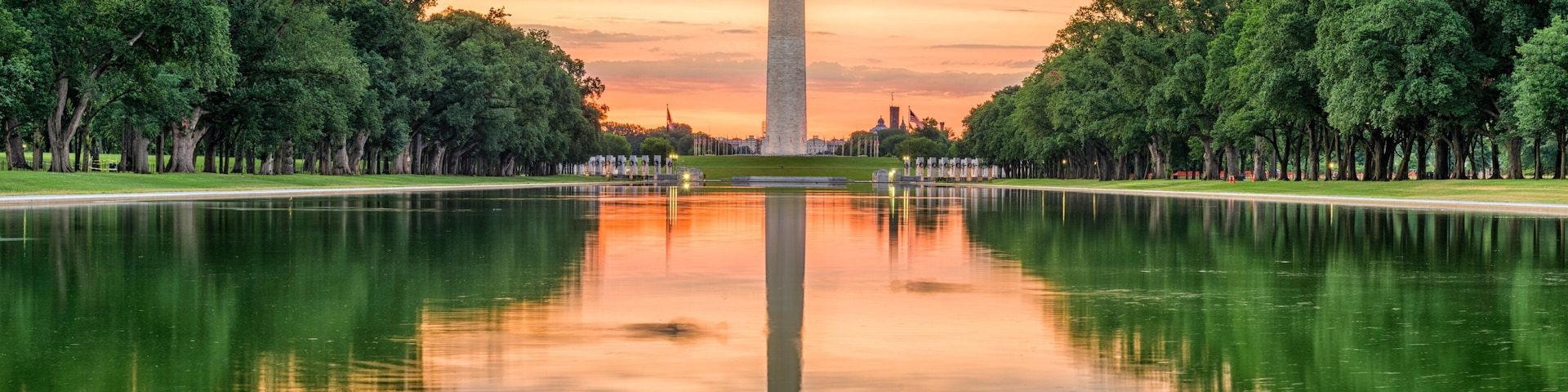 O Monumento a Washington refletido em águas calmas, rodeado por árvores e um céu ao pôr do sol.