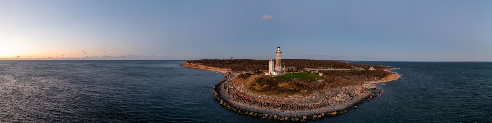 Vista aérea de um farol numa ilha rochosa, rodeada por um mar calmo.