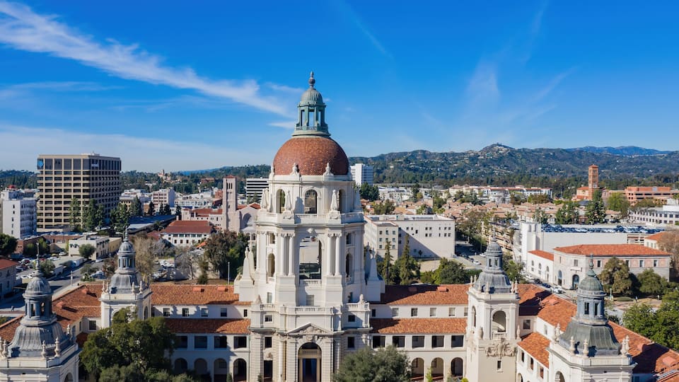Um edifício branco grande com uma cúpula e torres sineiras, cercado por edifícios com telhados vermelhos e árvores.